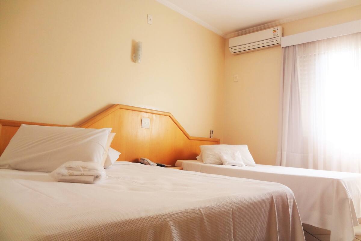 Quarto de hotel com duas camas, uma cabeceira de madeira, ar condicionado fixado na parede e uma janela com cortinas.