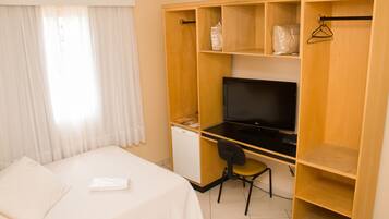 Quarto de hotel com uma cama, uma escrivaninha com televisão, um pequeno frigorífico e um guarda-roupa de madeira com prateleiras.