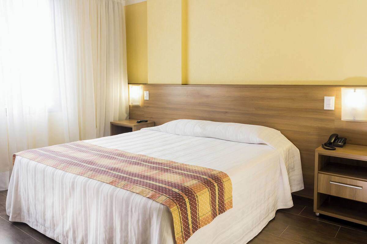 Quarto de hotel com uma cama grande, cabeceira de madeira, duas mesas de cabeceira com abajures e um telefone.