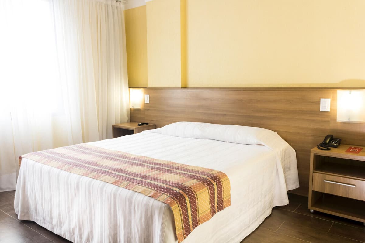 Quarto de hotel com uma cama grande, mesas de cabeceira, painel de cama de madeira e um criado-mudo com telefone.