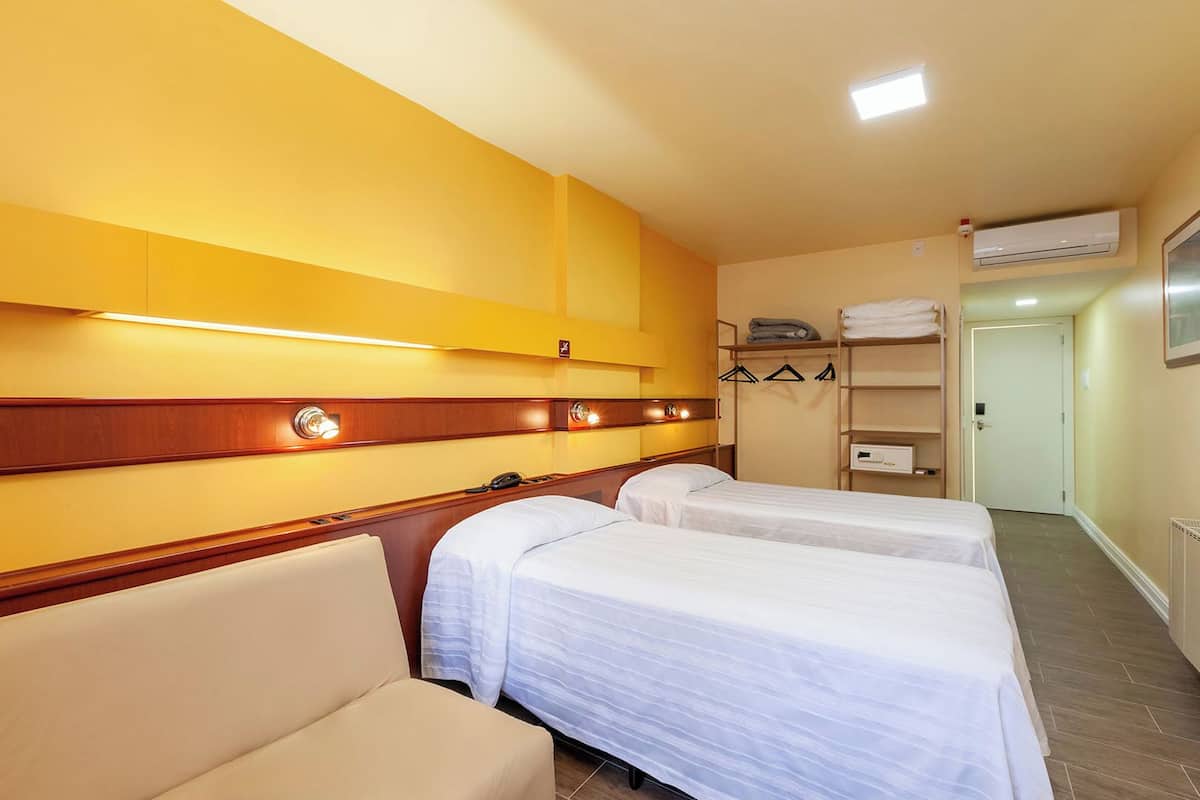 Quarto de hotel com duas camas, cabeceira de madeira, mesa de cabeceira, guarda-roupa e ar condicionado.