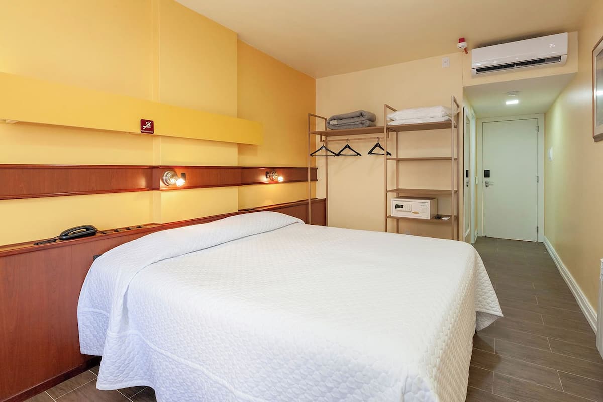 Quarto de hotel com uma cama, cabeceira de madeira, mesa de cabeceira com telefone, aquecedor fixado na parede, prateleira com toalhas e unidade de ar condicionado.