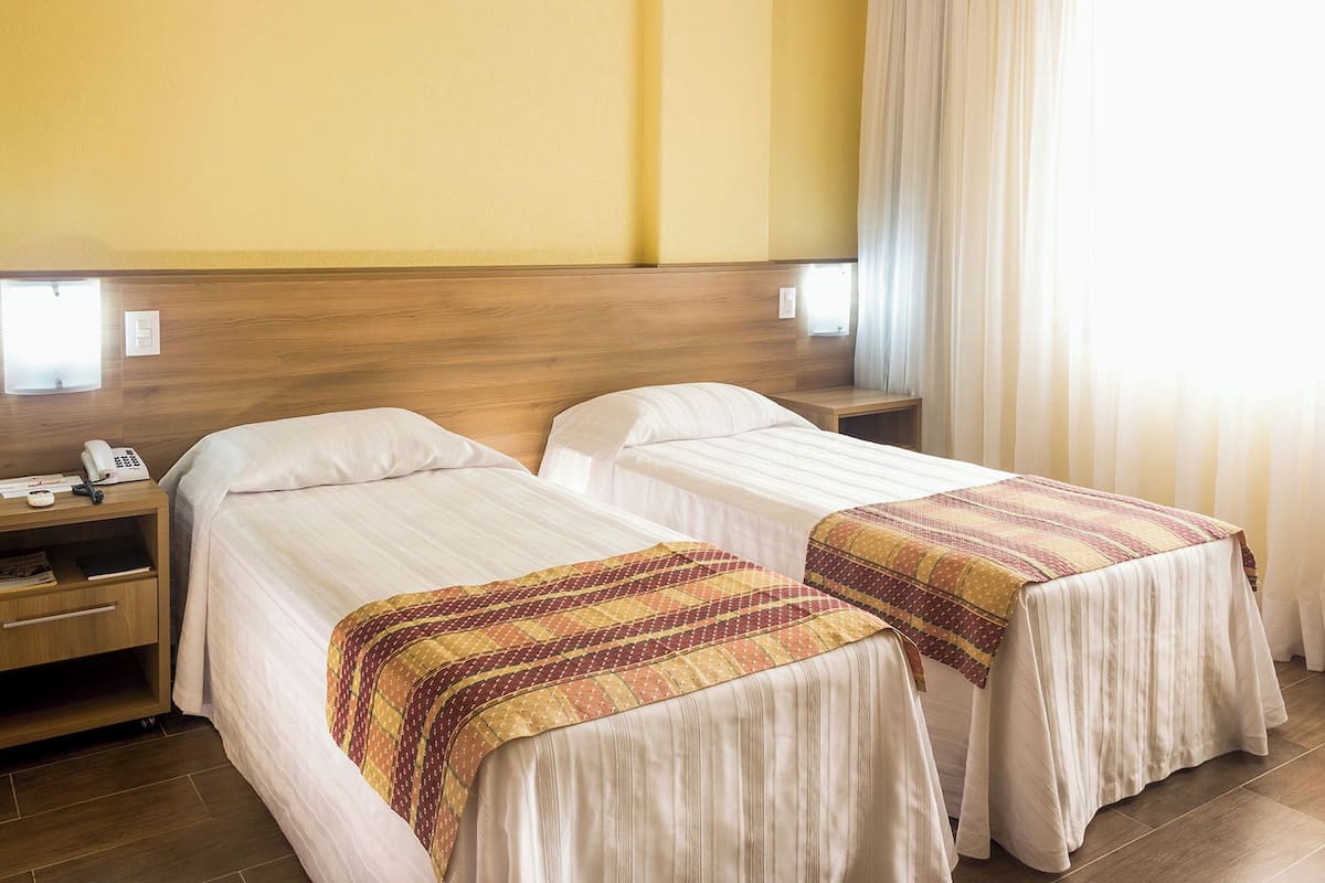 Quarto de hotel com duas camas, cabeceira de madeira, mesas de cabeceira com telefone e abajur, e uma janela com cortinas.