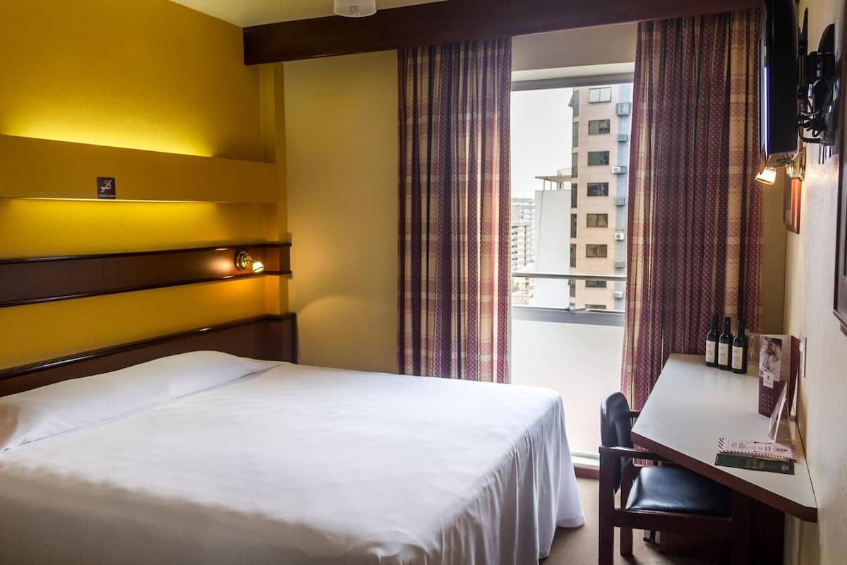 Quarto de hotel com cama, escrivaninha e vista para edifícios através de cortinas.