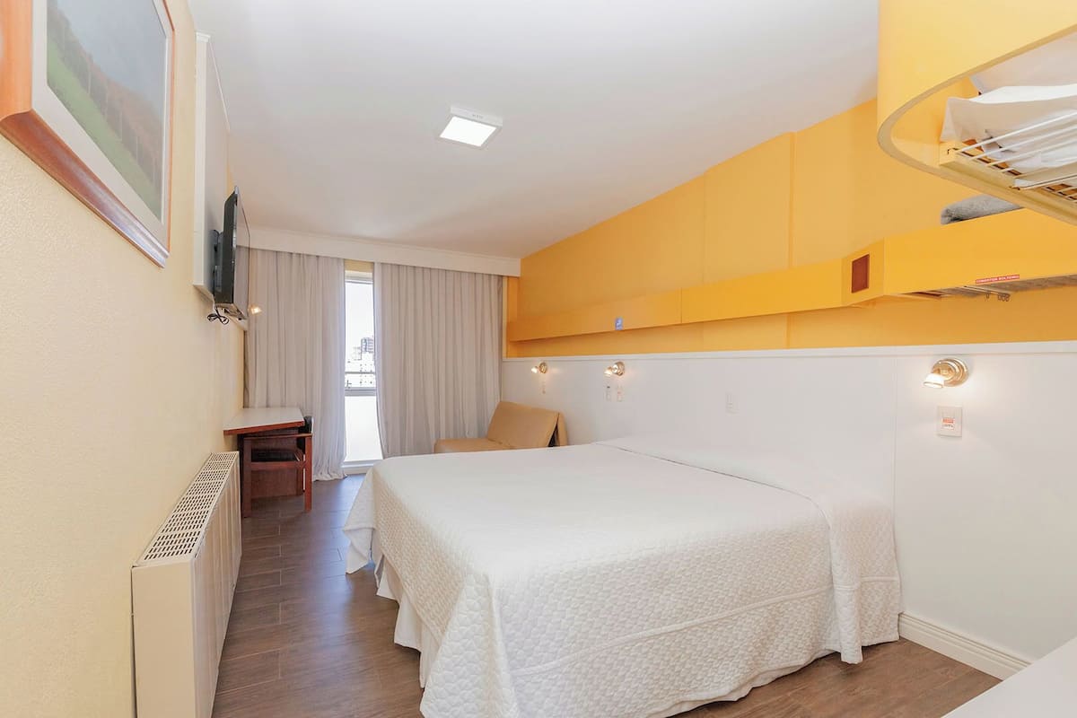 Quarto de hotel com cama, escrivaninha, cadeira, radiador e janela com cortinas.