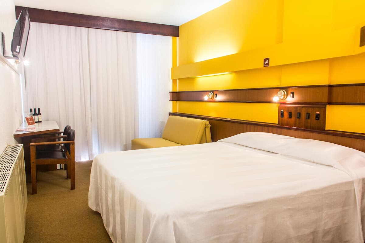 Quarto de hotel com uma cama, uma escrivaninha com cadeira, uma mesinha com garrafas e uma janela com cortinas.