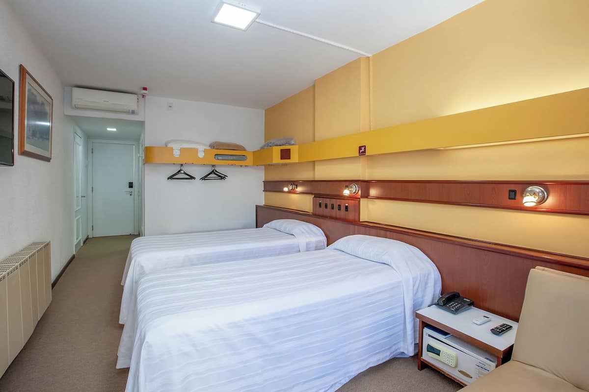 Quarto de hotel com duas camas, uma escrivaninha com telefone e controles remotos, e um corredor que dá para outro quarto.
