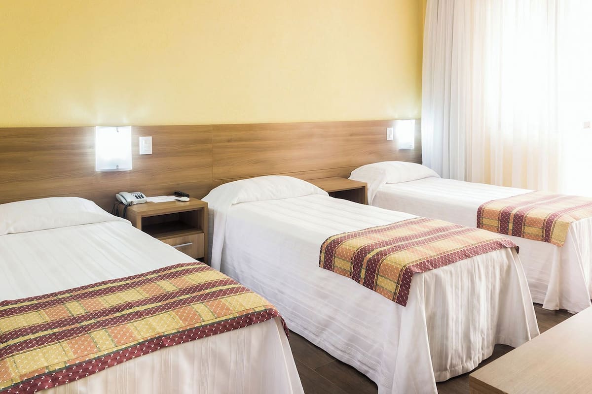 Quarto de hotel com três camas de solteiro, cada uma com colcha estampada. Possui cabeceira de madeira, mesa de cabeceira com telefone e janela com cortinas.