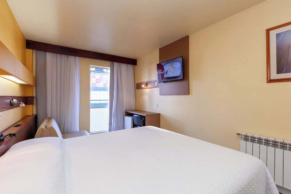 Quarto de hotel com cama, escrivaninha, cadeira, televisão e janela com cortinas.