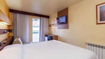 Quarto de hotel com cama, escrivaninha, cadeira, televisão e janela com cortinas.