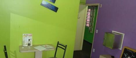 Um quarto com uma parede verde, uma parede roxa, uma mesa com cadeiras, um cartaz na parede e uma porta que dá para outro quarto.
