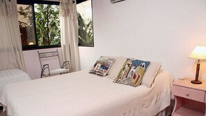 Quarto com cama, duas almofadas, mesa de cabeceira, abajur e janela com cortinas.