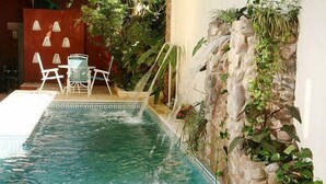 Piscina com cascata, cercada por um muro de pedra e vegetação. Há uma mesa com cadeiras ao fundo.