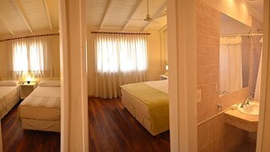 Quarto com duas camas, piso de madeira, banheiro com pia e espelho, e janela com cortinas.
