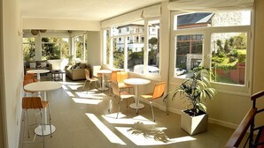 Um quarto claro e ensolarado, com grandes janelas, um sofá e várias cadeiras dispostas em torno de uma mesa central.