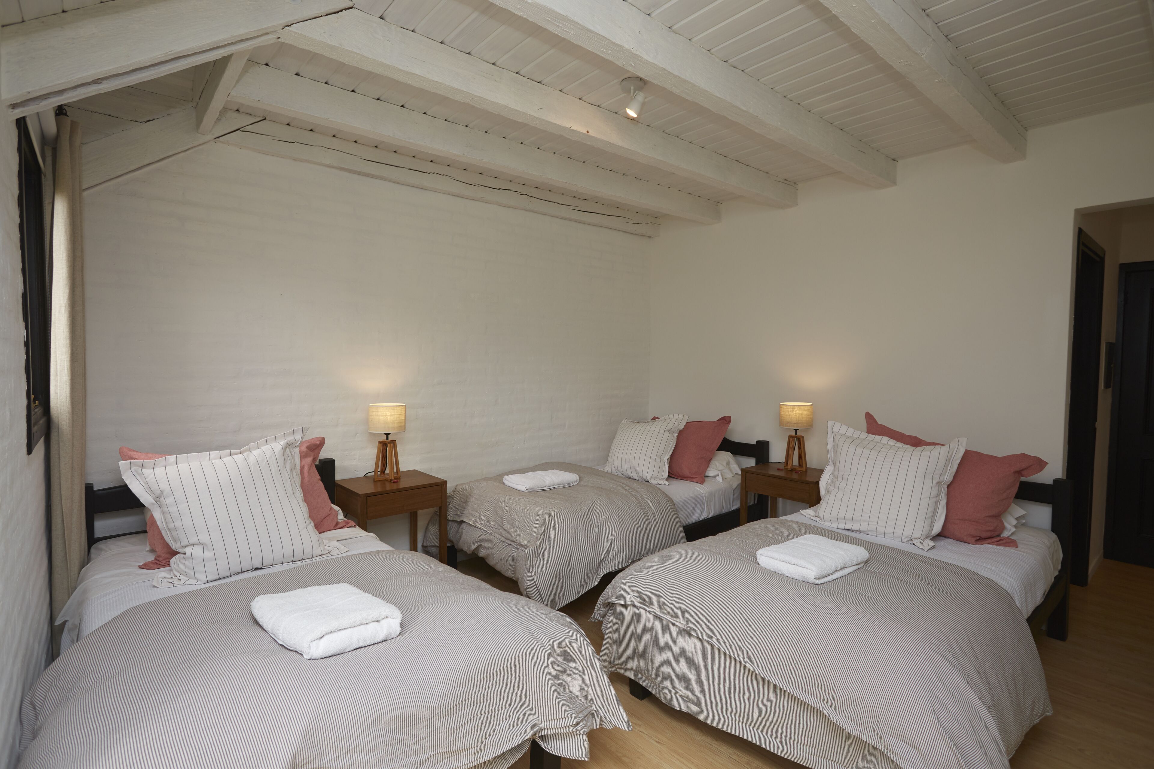 Quarto de hotel com três camas de solteiro, cada uma com travesseiro e mesa de cabeceira com abajur.