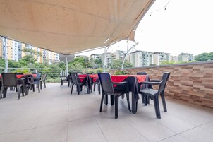 Uma área de refeições ao ar livre com mesas e cadeiras, um espaço coberto e vista para edifícios residenciais ao fundo.