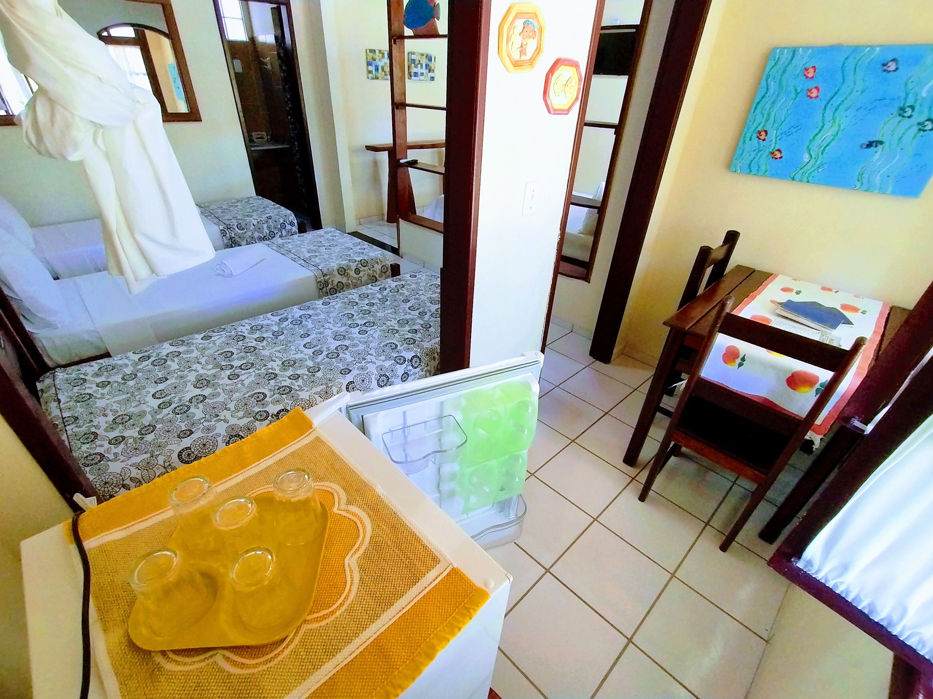 Quarto de hotel com duas camas, uma mesa pequena e uma cadeira. Há uma janela com cortinas, um espelho e uma parede com pratos decorativos.