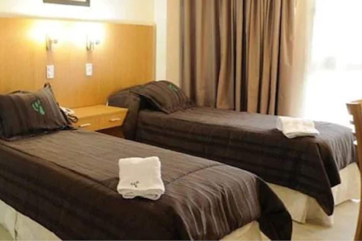 Quarto de hotel com duas camas, uma cabeceira de madeira, uma mesinha de cabeceira e uma cadeira.