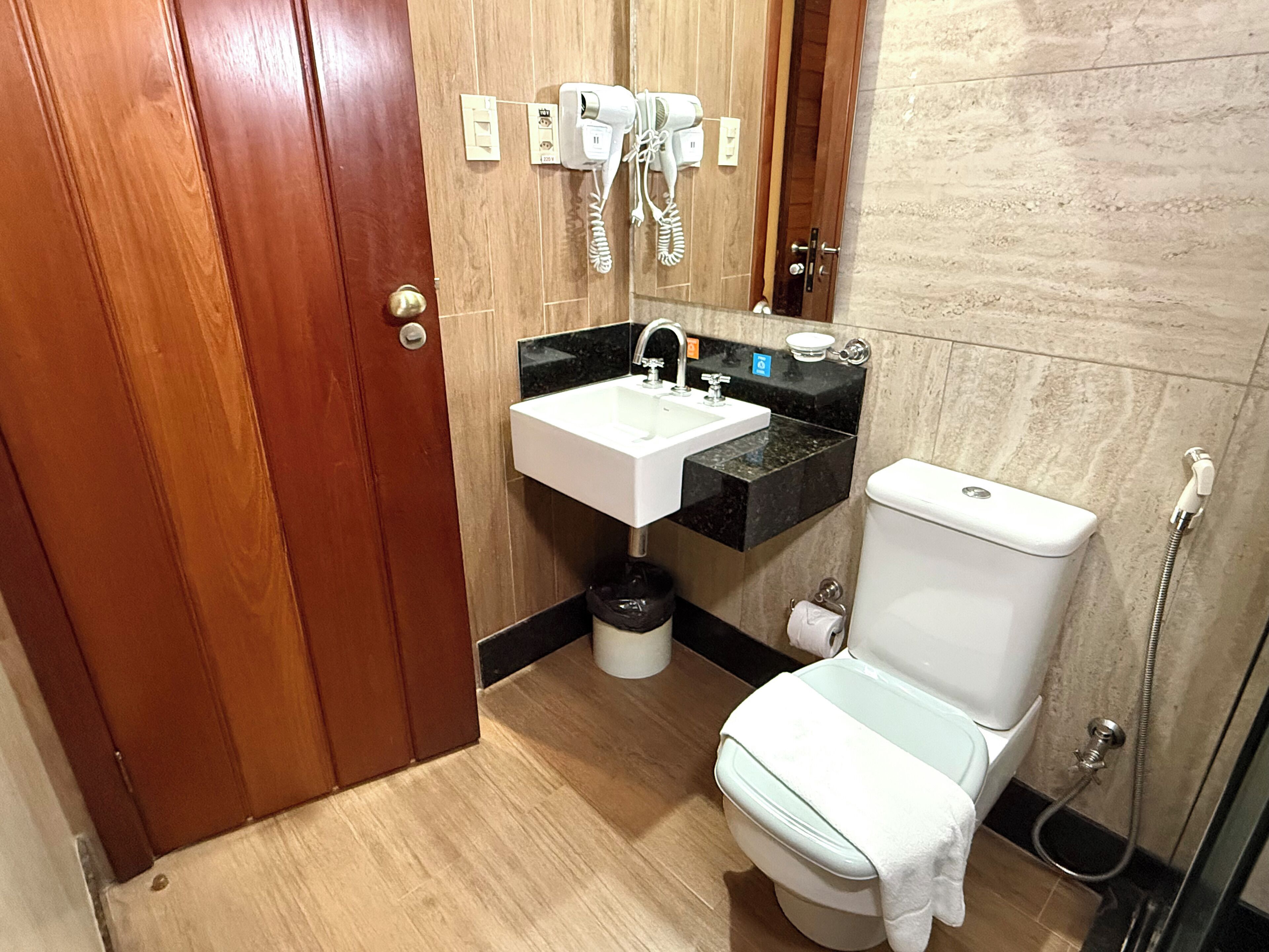 Casa de banho compacta com porta de madeira, lavatório branco com bancada preta, sanita e zona de duche.