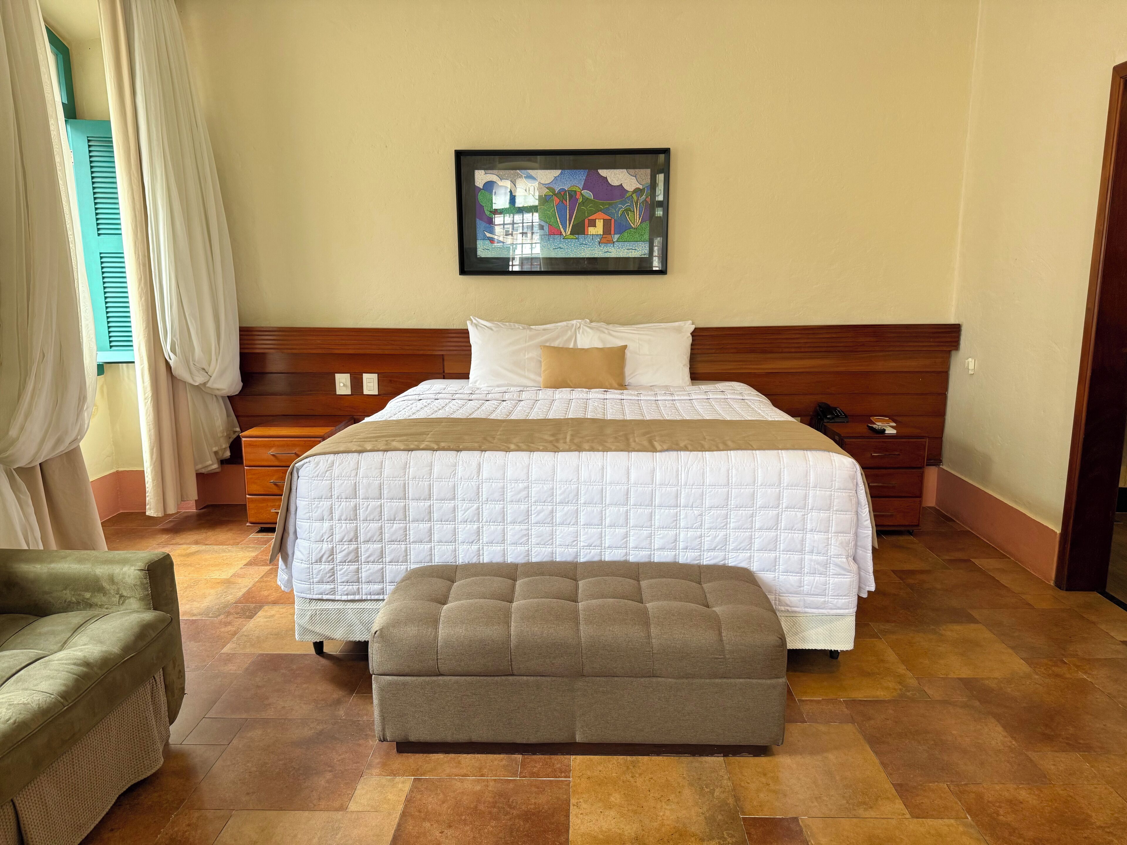 Quarto de hotel com uma cama grande, cabeceira de madeira, mesa de cabeceira, poltrona verde e uma moldura com um quadro na parede.