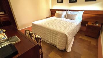 Quarto de hotel com uma cama grande, cabeceira de madeira, mesa de cabeceira com abajur e uma cadeira.