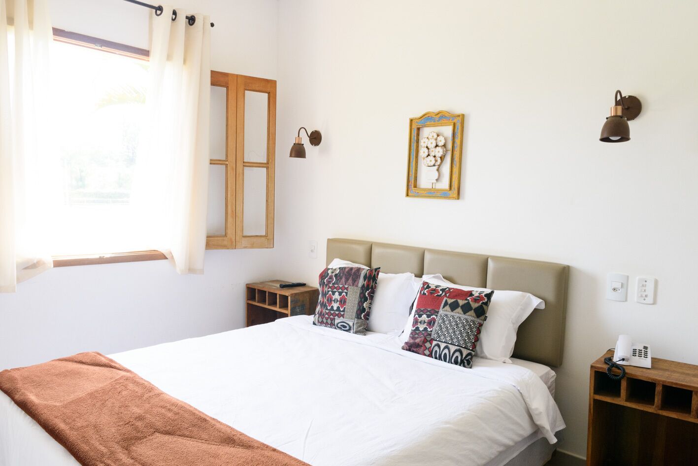 Cama bem arrumada com almofadas estampadas, mesa de cabeceira com telefone, luminária na parede e janela com cortinas.