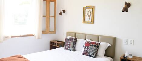 Cama bem arrumada com almofadas estampadas, mesa de cabeceira com telefone, luminária na parede e janela com cortinas.