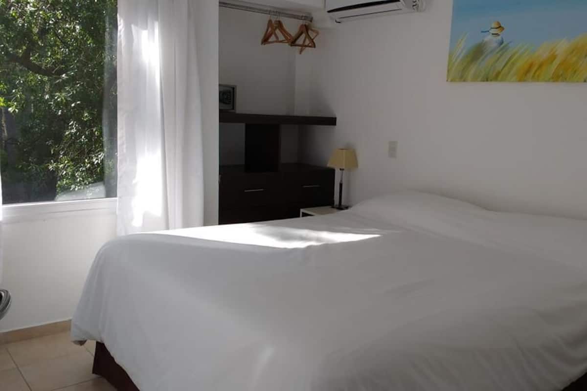 Quarto com cama, mesa de cabeceira, abajur e um quadro na parede.