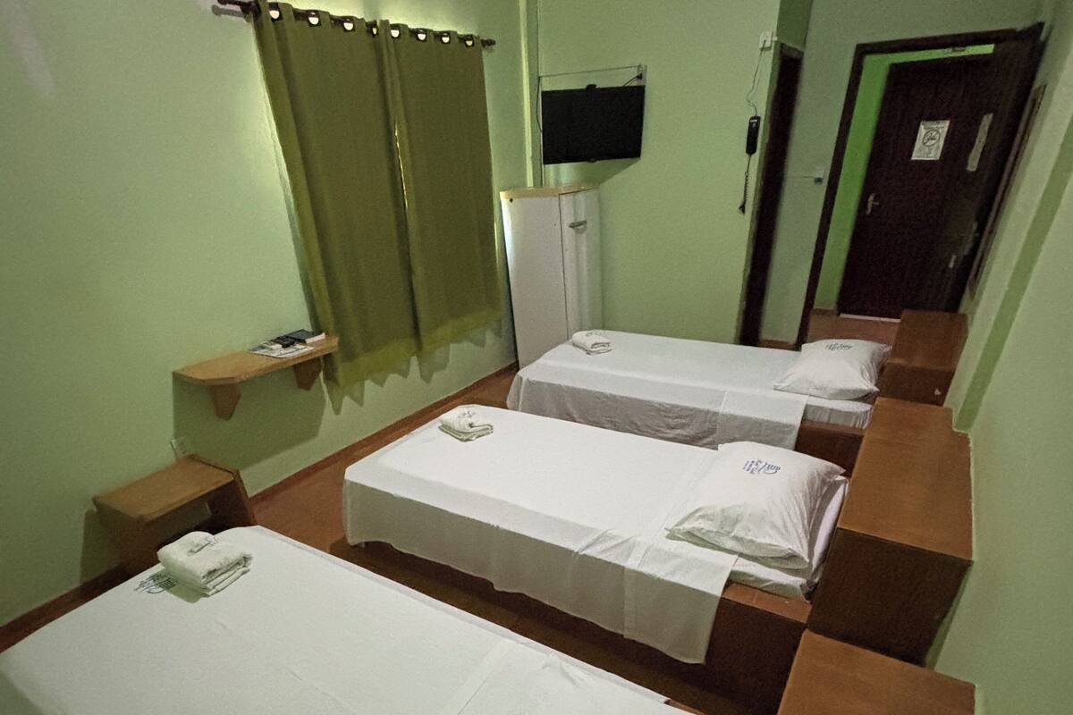 Quarto de hotel com duas camas de solteiro, uma pequena mesa, um prateleira e uma televisão na parede.