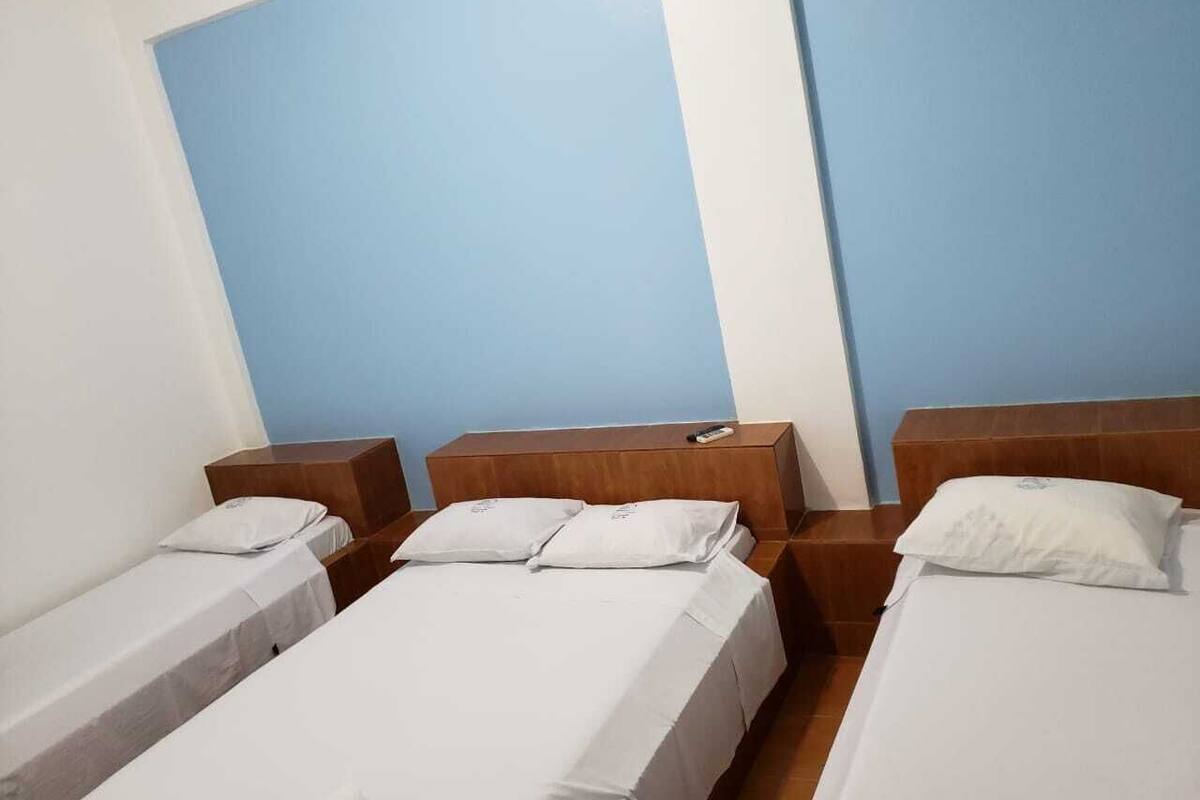 Quarto de hotel com três camas de solteiro, cada uma com cabeceira de madeira e roupa de cama branca.