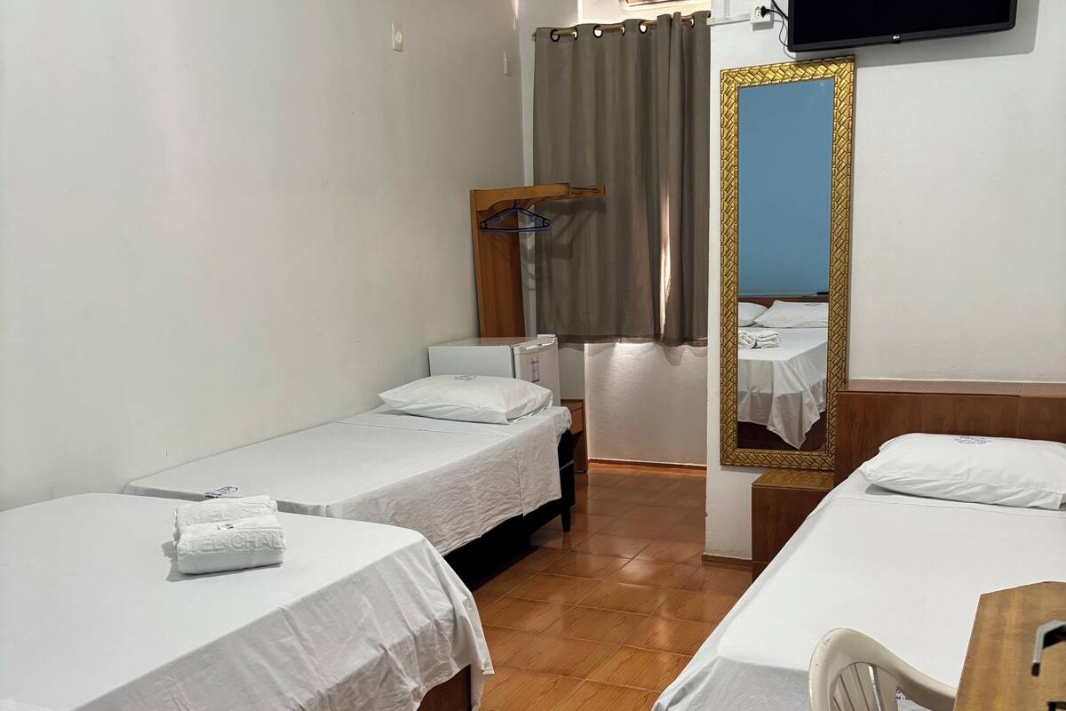 Quarto de hotel com duas camas de solteiro, uma cadeira de madeira, uma televisão na parede e um espelho.