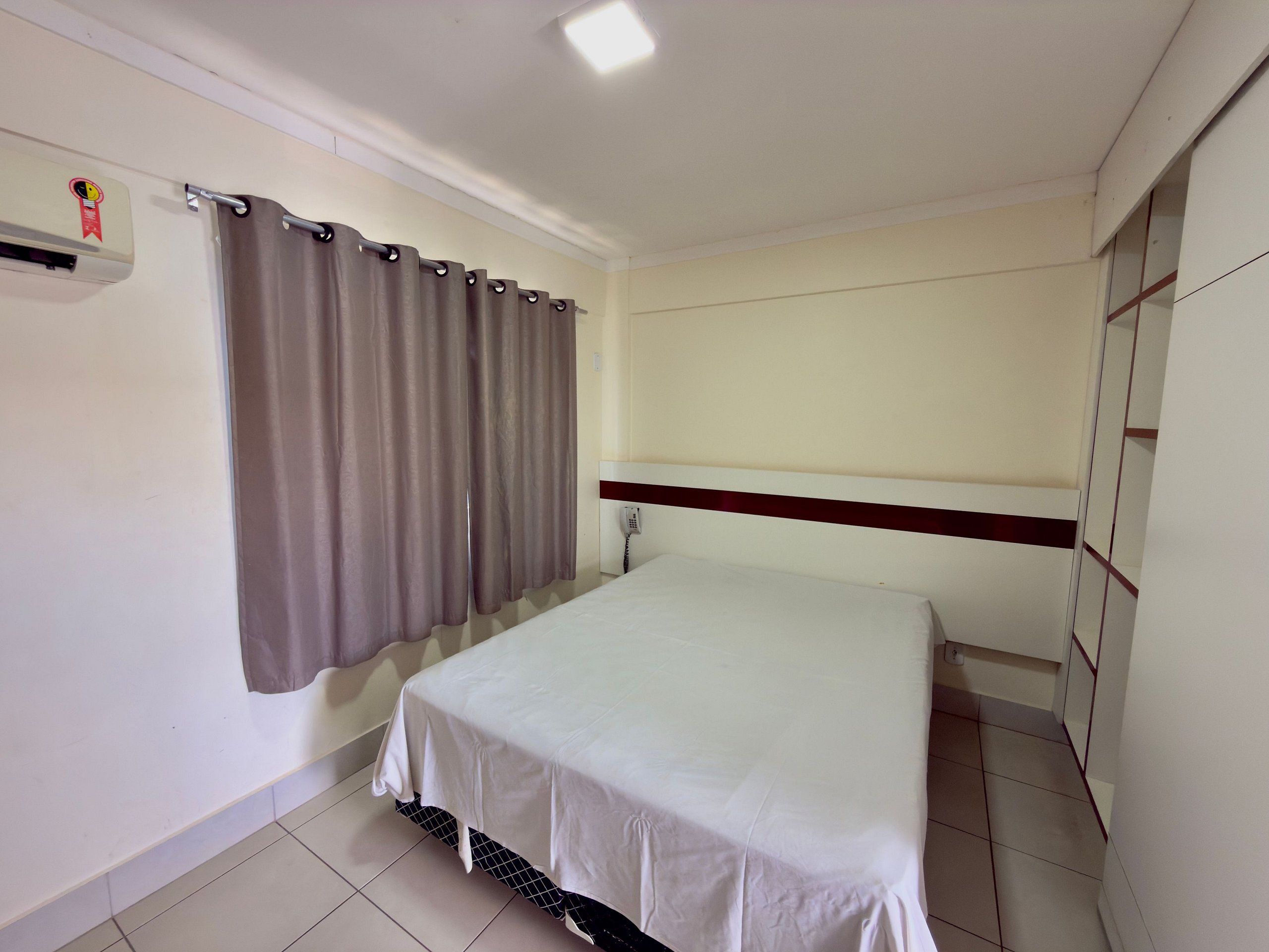 Quarto de hotel com cama, cortinas, telefone e ar condicionado fixado na parede.