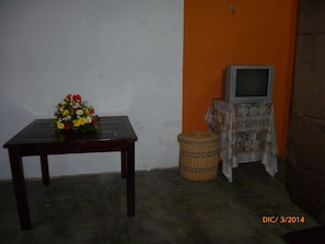 Quarto com uma mesa de madeira, um banquinho de vime, uma televisão sobre um suporte e um arranjo floral.