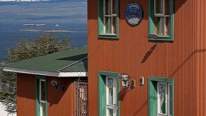 Uma casa de madeira vermelha com detalhes verdes, um emblema azul na parede e vista para o oceano.