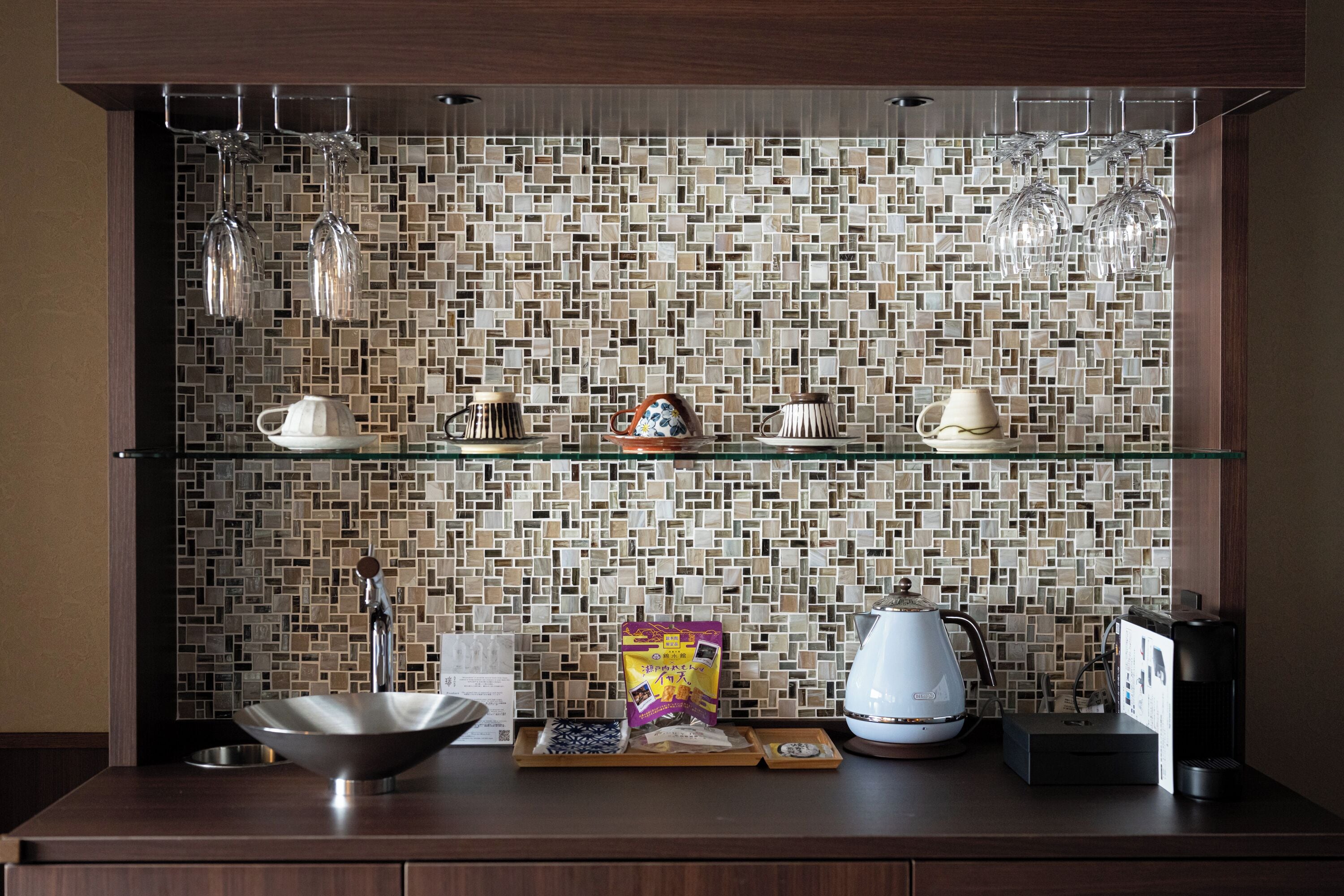Uma cozinha com painel de azulejos, pia de aço inoxidável, cafeteira e bandeja com embalagens de lanches.