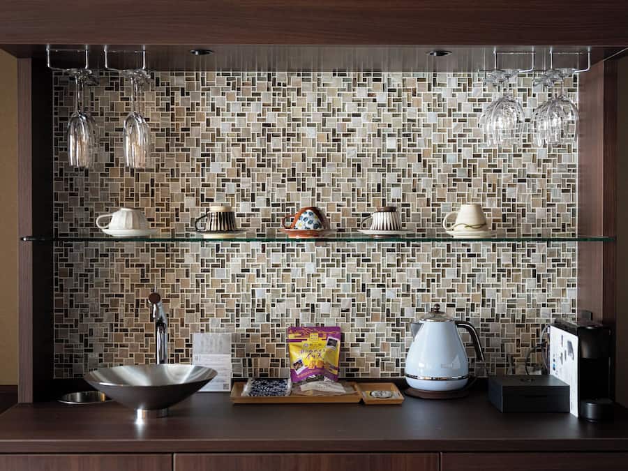 Uma cozinha com painel de azulejos, pia de aço inoxidável, cafeteira e bandeja com embalagens de lanches.