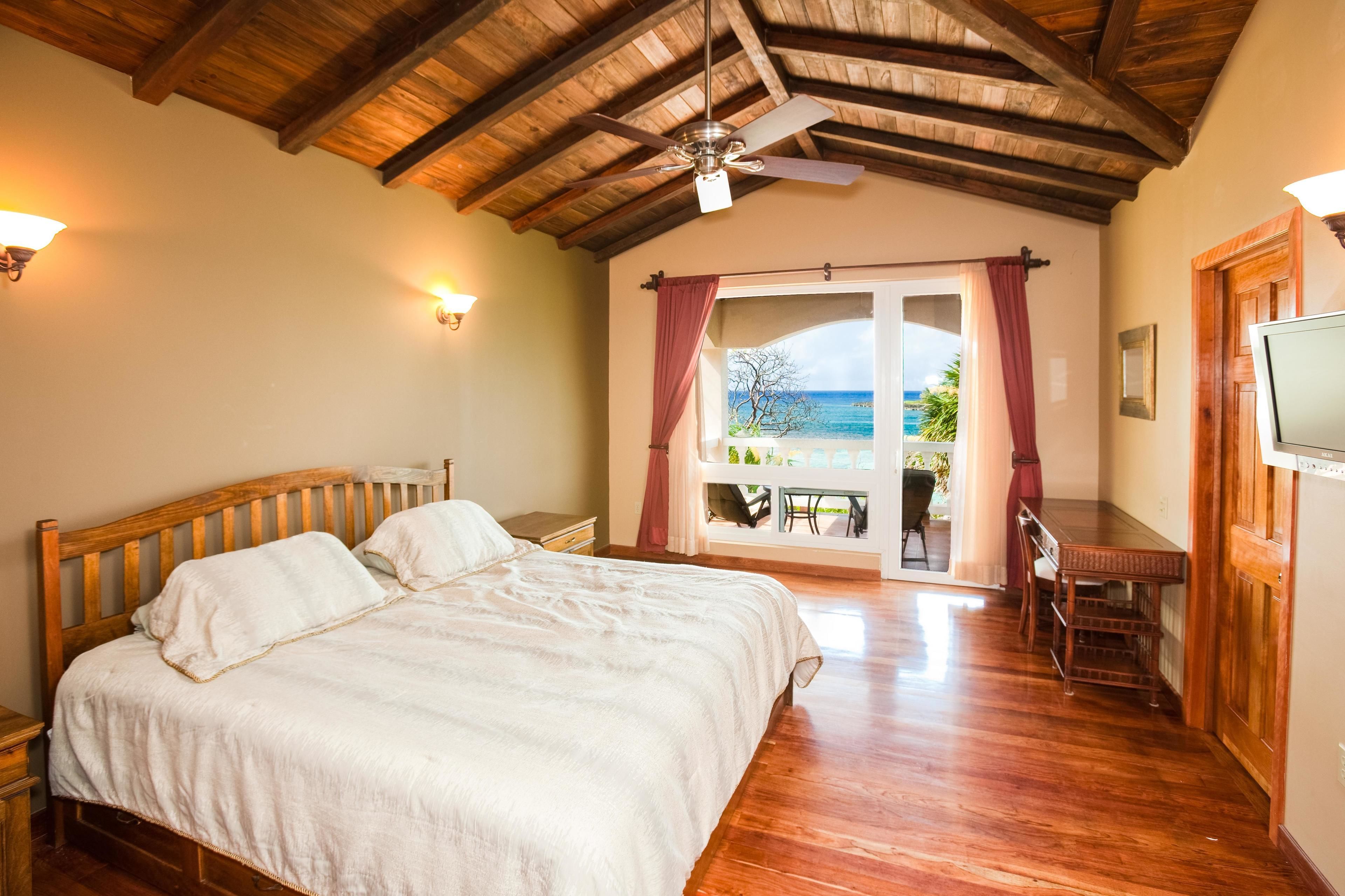 Quarto com teto de madeira, uma cama, uma escrivaninha e vista para o mar pela janela.