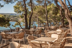 Uma área de refeições ao ar livre com móveis de vime e vista para um lago e colinas.