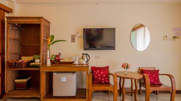 Quarto de hotel com secretária de madeira, televisão, mini-frigorífico e área de estar com duas cadeiras e uma mesa.