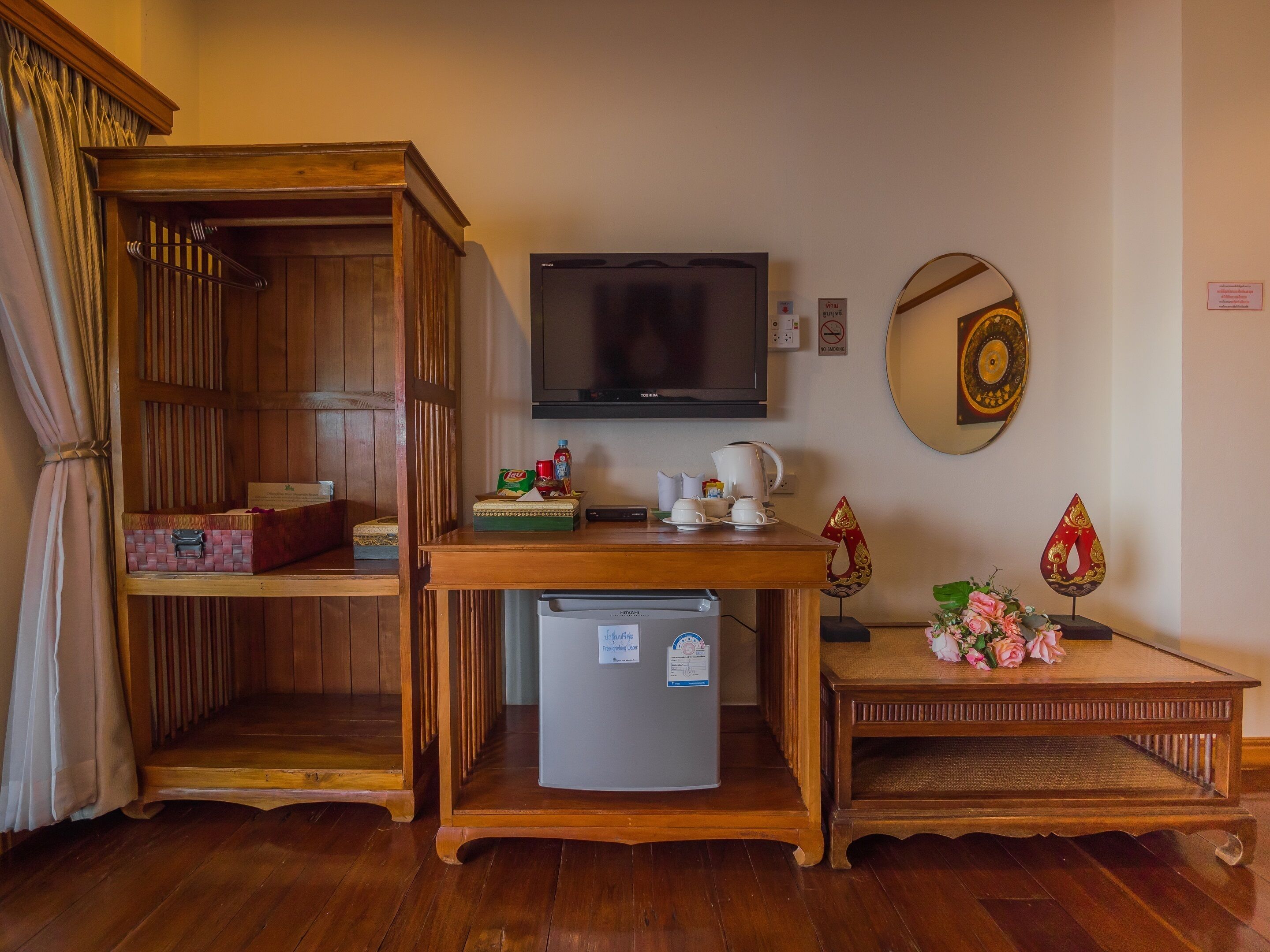 Quarto com armário de madeira, televisão na parede, pequena geladeira e mesa com bule e xícaras.
