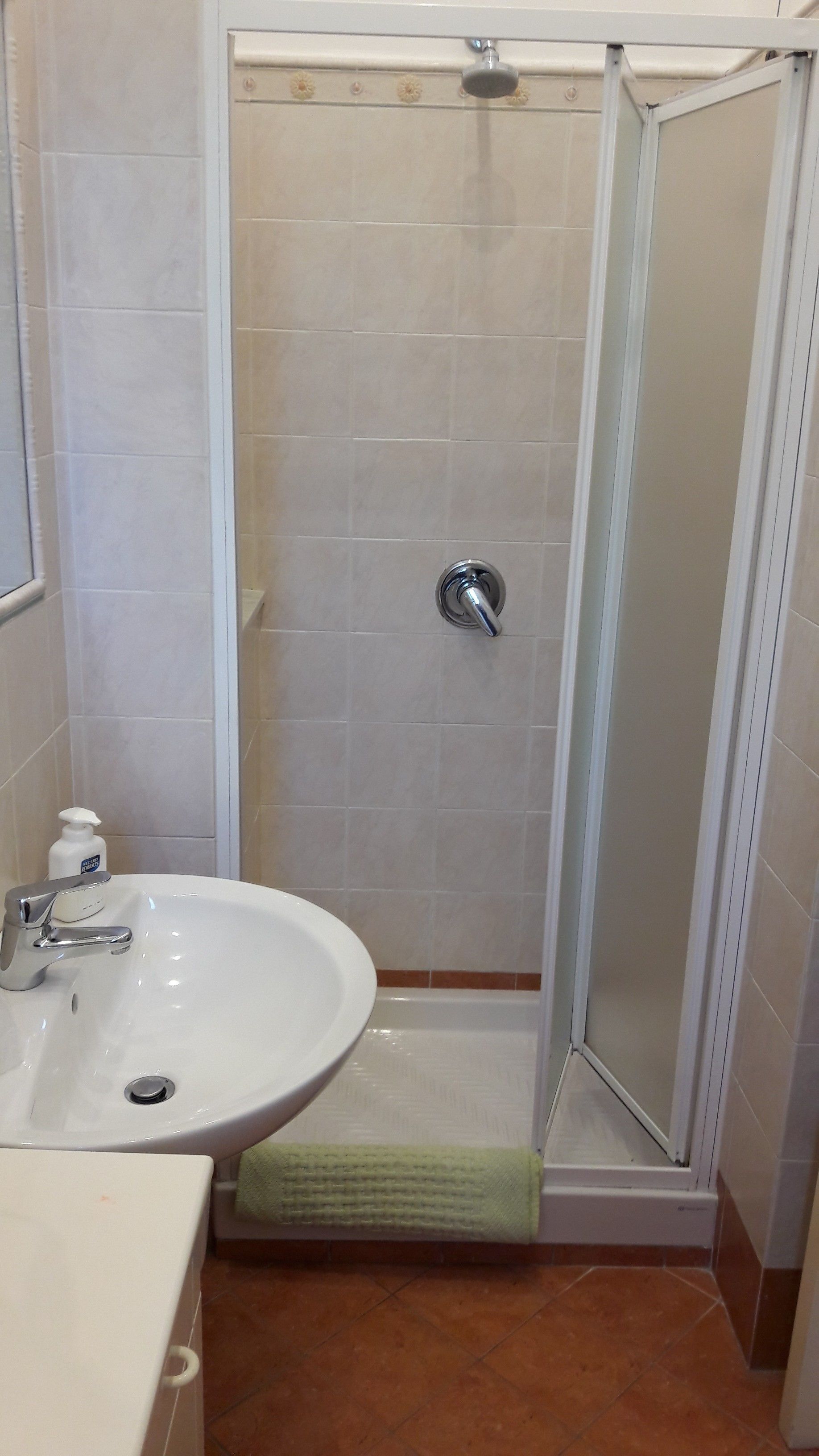 Casa de banho com lavatório branco, chuveiro com porta de vidro e toalha verde no chão.