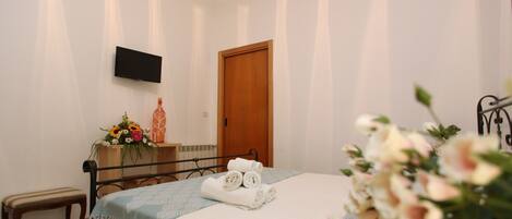 Um quarto com uma cama, uma mesinha de cabeceira com um vaso de flores, um banco de madeira e uma televisão na parede.