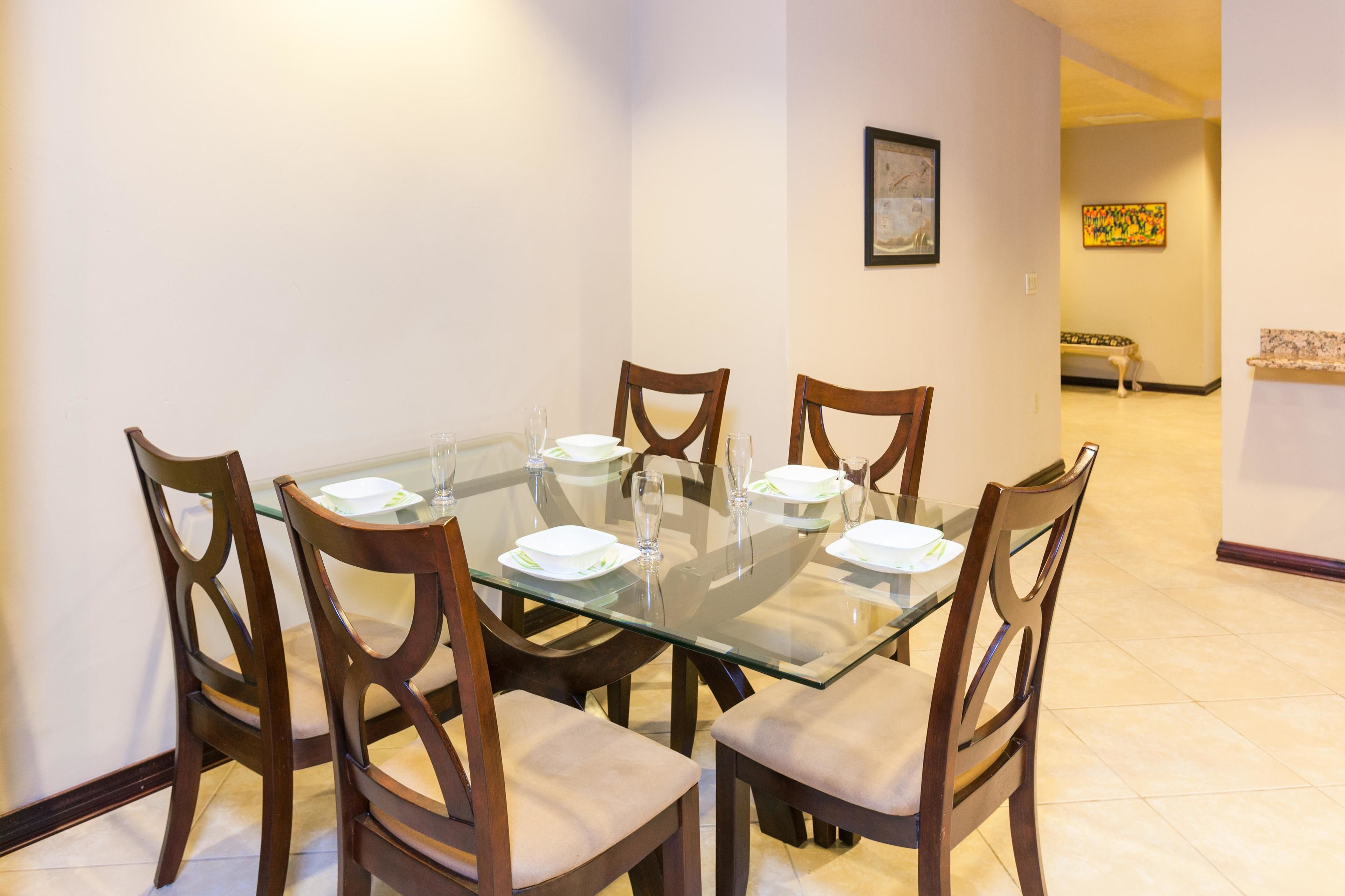 Uma área de refeições com mesa de vidro e cadeiras de madeira, um quadro na parede e um banco ao fundo.