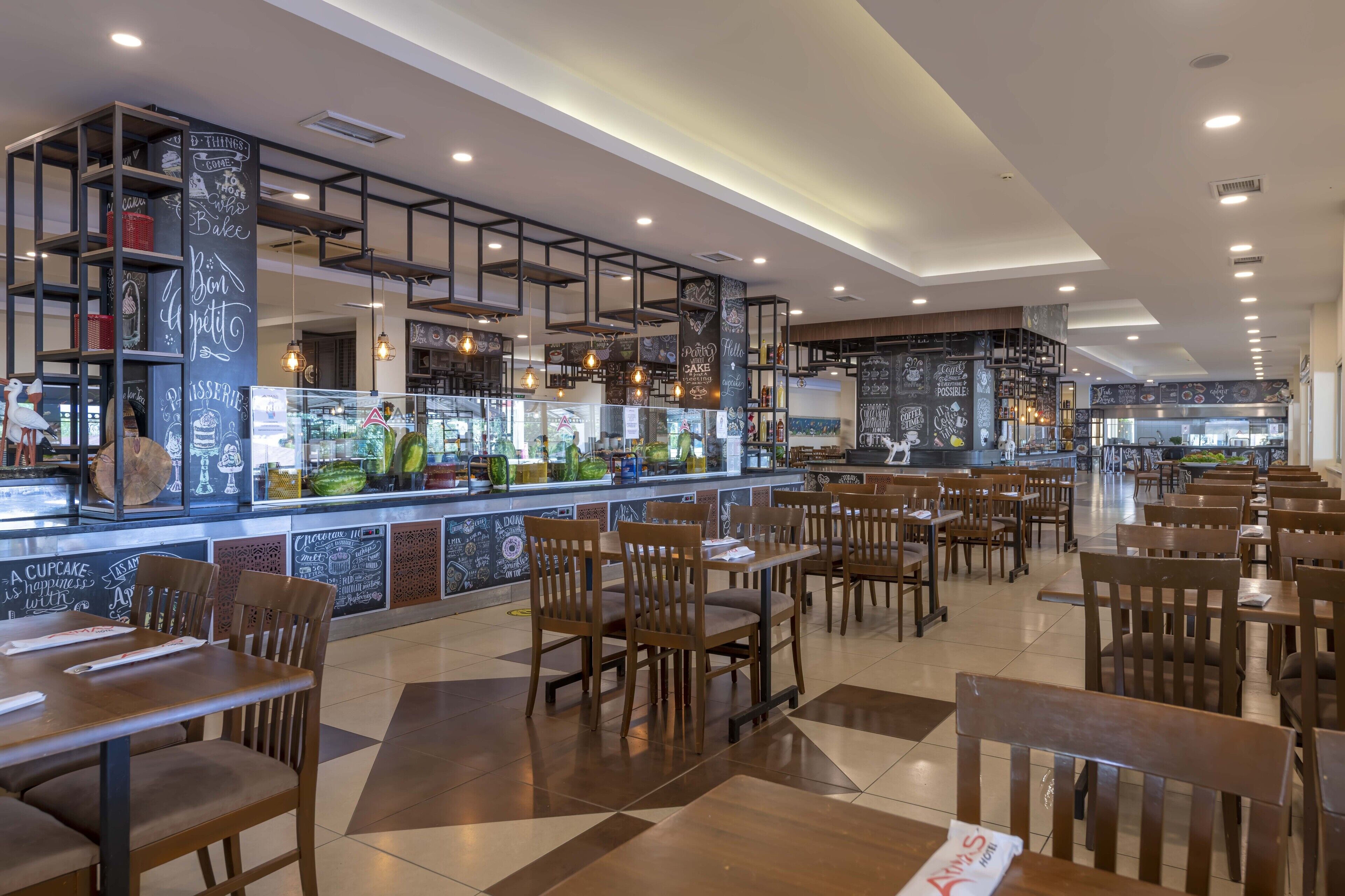 Um café moderno com mesas e cadeiras de madeira, um cardápio em lousa e diversos alimentos expostos ao fundo.