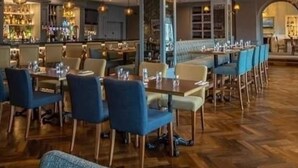 Restaurante com piso de madeira, cadeiras azuis e bege e um espaço de bar com balcão.