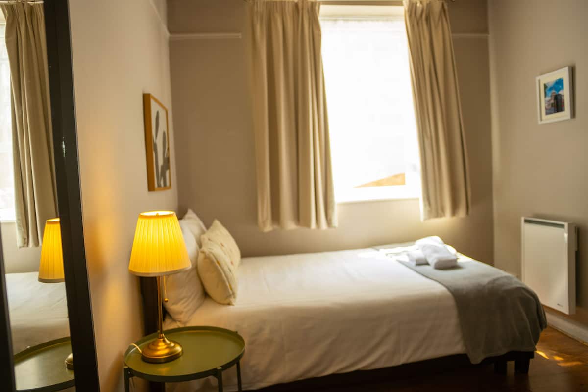 Cama bem arrumada com edredom branco e almofadas, mesa de cabeceira com abajur e janela com cortinas.