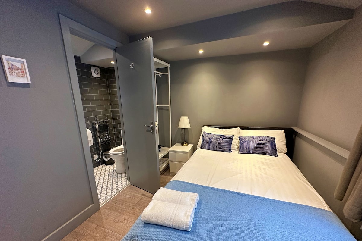 Quarto de hotel com uma cama, mesa de cabeceira, luminária e banheiro visível por uma porta aberta.