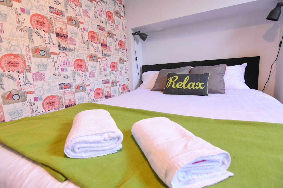 Cama bem arrumada com colcha verde, duas toalhas enroladas e um travesseiro com a palavra 'Relax'.