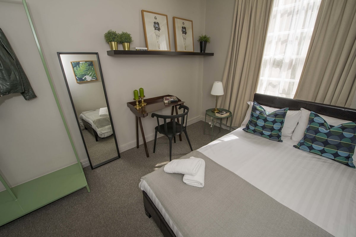 Quarto de hotel com cama, escrivaninha, cadeira, espelho, mesa pequena com abajur e prateleira com plantas e quadros.